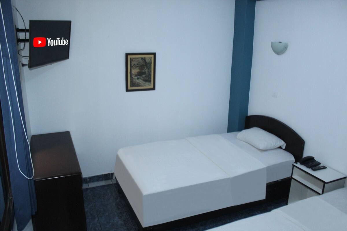 Quarto de hotel com uma cama, uma mesa de cabeceira com abajur, uma escrivaninha com computador e o logotipo do YouTube na parede.