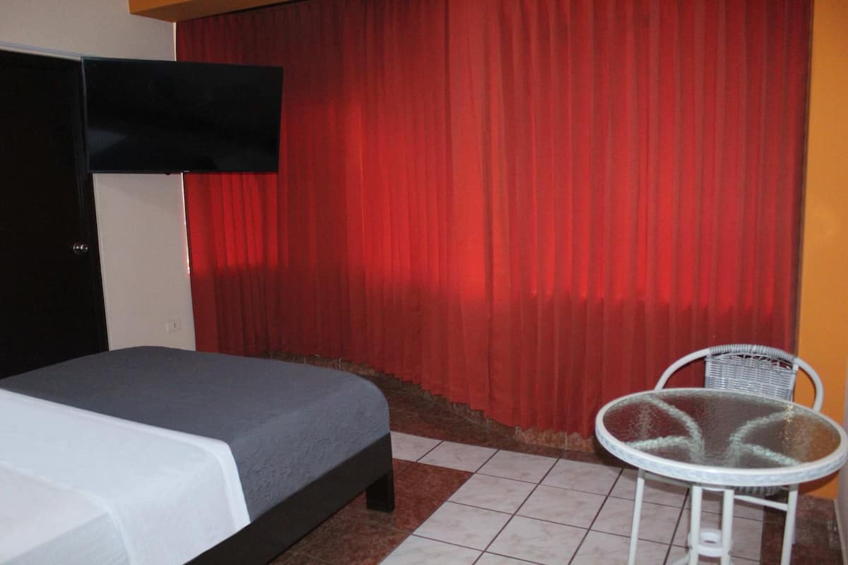 Quarto de hotel com uma cama, televisão na parede, cortina vermelha, mesa redonda pequena e uma cadeira.