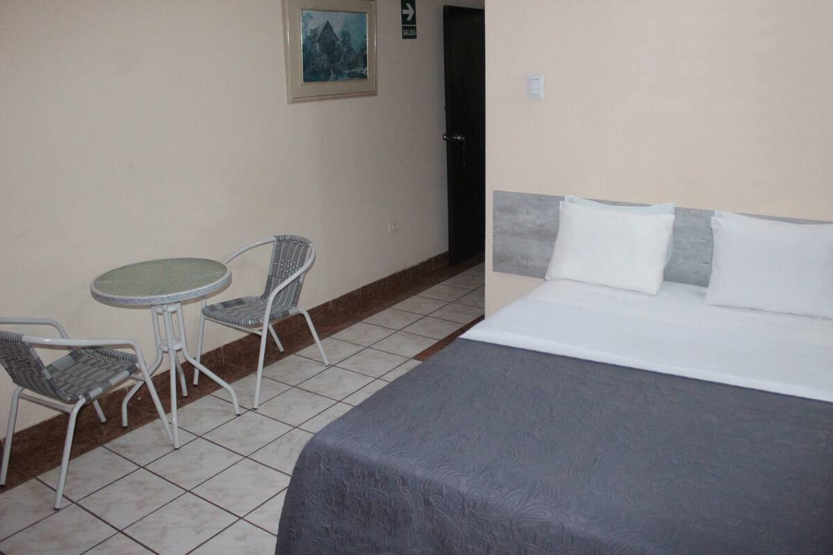 Quarto de hotel com cama, duas cadeiras, uma mesinha e um quadro na parede.