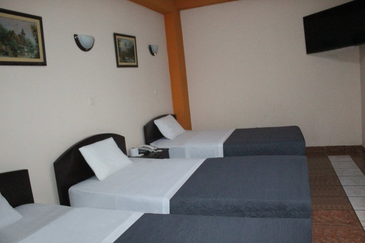 Quarto de hotel com três camas de solteiro, uma pequena mesa e uma televisão presa à parede.