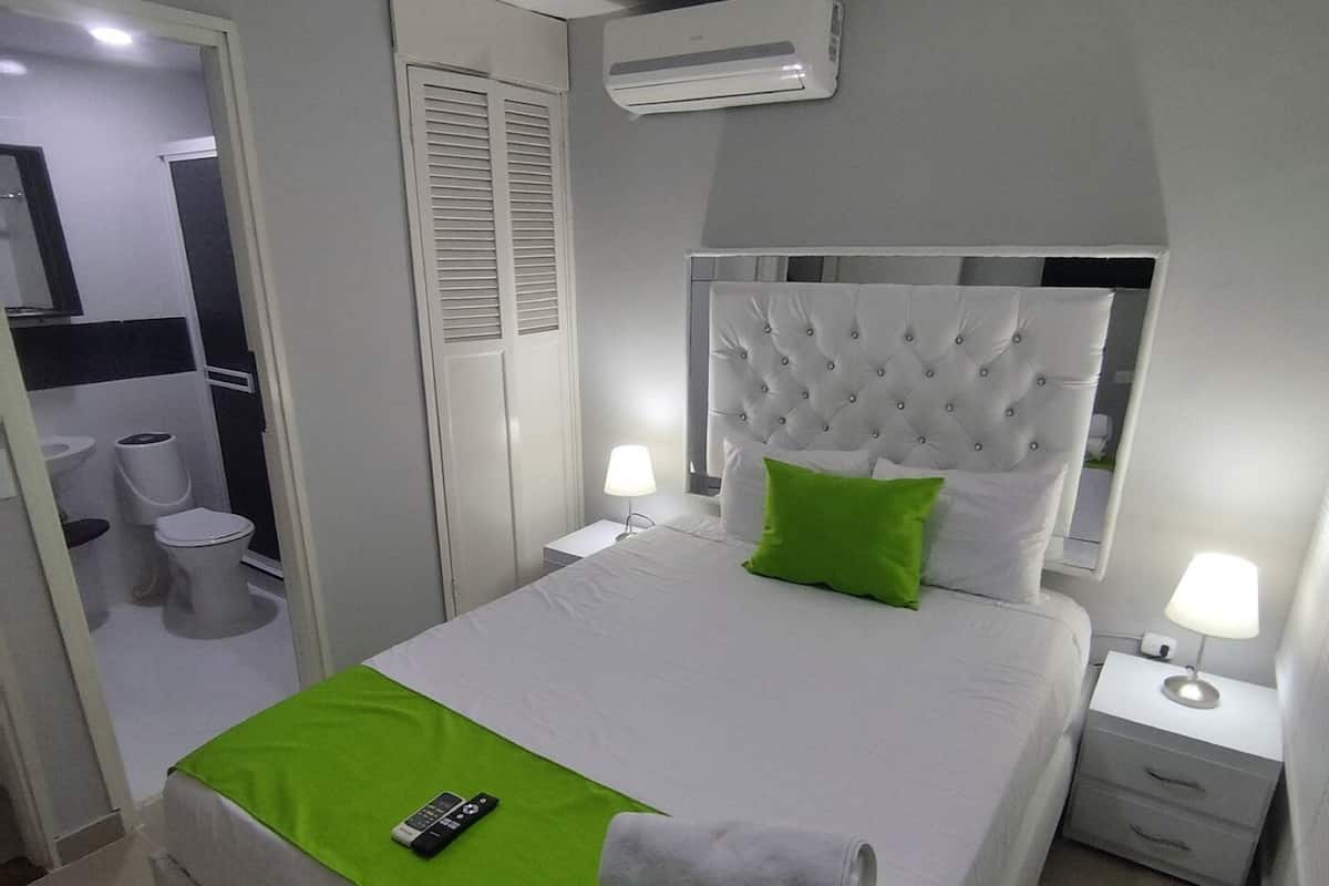 Quarto bem arrumado com uma cama grande, uma mesa de cabeceira, um ar condicionado e um banheiro visível através de uma porta aberta.
