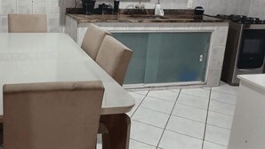 Uma área de refeições com uma mesa retangular e quatro cadeiras, uma cozinha com fogão e pia, e um chão revestido de azulejos.