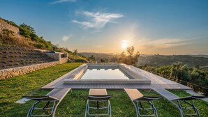 Uma piscina retangular com borda de pedra, cercada por grama e uma parede de pedra. Quatro cadeiras de descanso estão dispostas em frente à piscina.