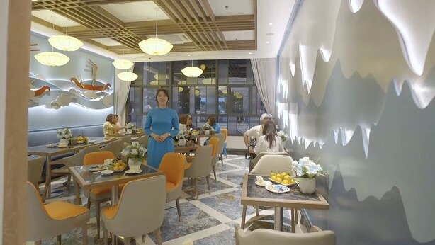 Um restaurante com mesas e cadeiras, uma mulher de vestido azul e uma parede com um desenho de montanha.