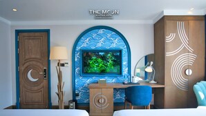 Quarto de hotel com televisão, escrivaninha, duas camas e um detalhe decorativo na parede.