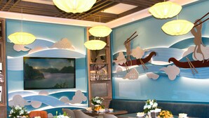 Interior de restaurante com tema asiático tradicional, apresentando um grande mural de um barco e ondas, vários lanternas penduradas e uma tela de televisão exibindo uma vista panorâmica.