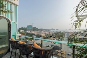 Uma varanda com cadeiras de vime e guarda-corpo de vidro, com vista para um horizonte urbano com edifícios e uma ponte.