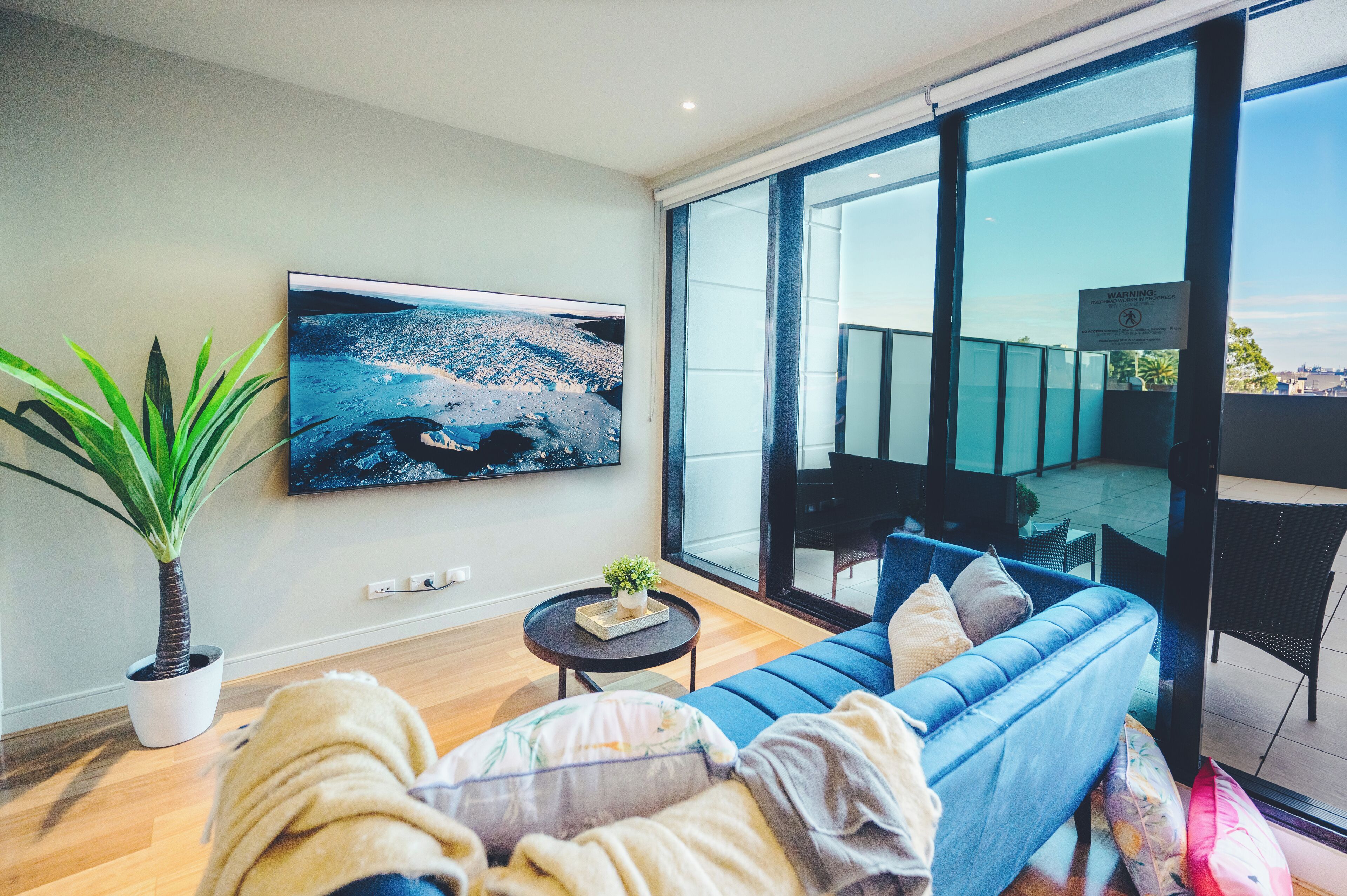 Uma sala de estar moderna com uma televisão de tela plana grande, um sofá azul, uma mesinha de centro redonda e um vaso de planta.