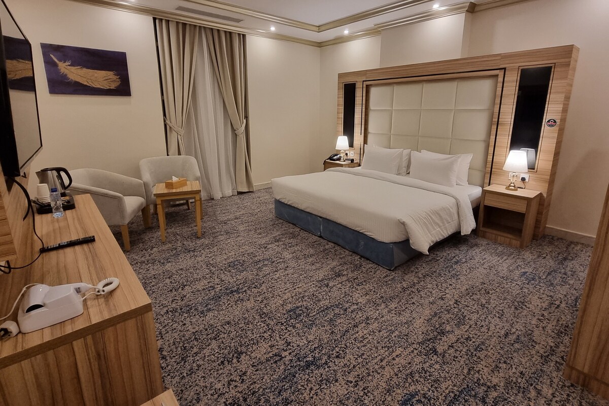 Quarto de hotel com uma cama grande, duas mesas de cabeceira, uma escrivaninha com telefone, uma cadeira e piso coberto de carpete.