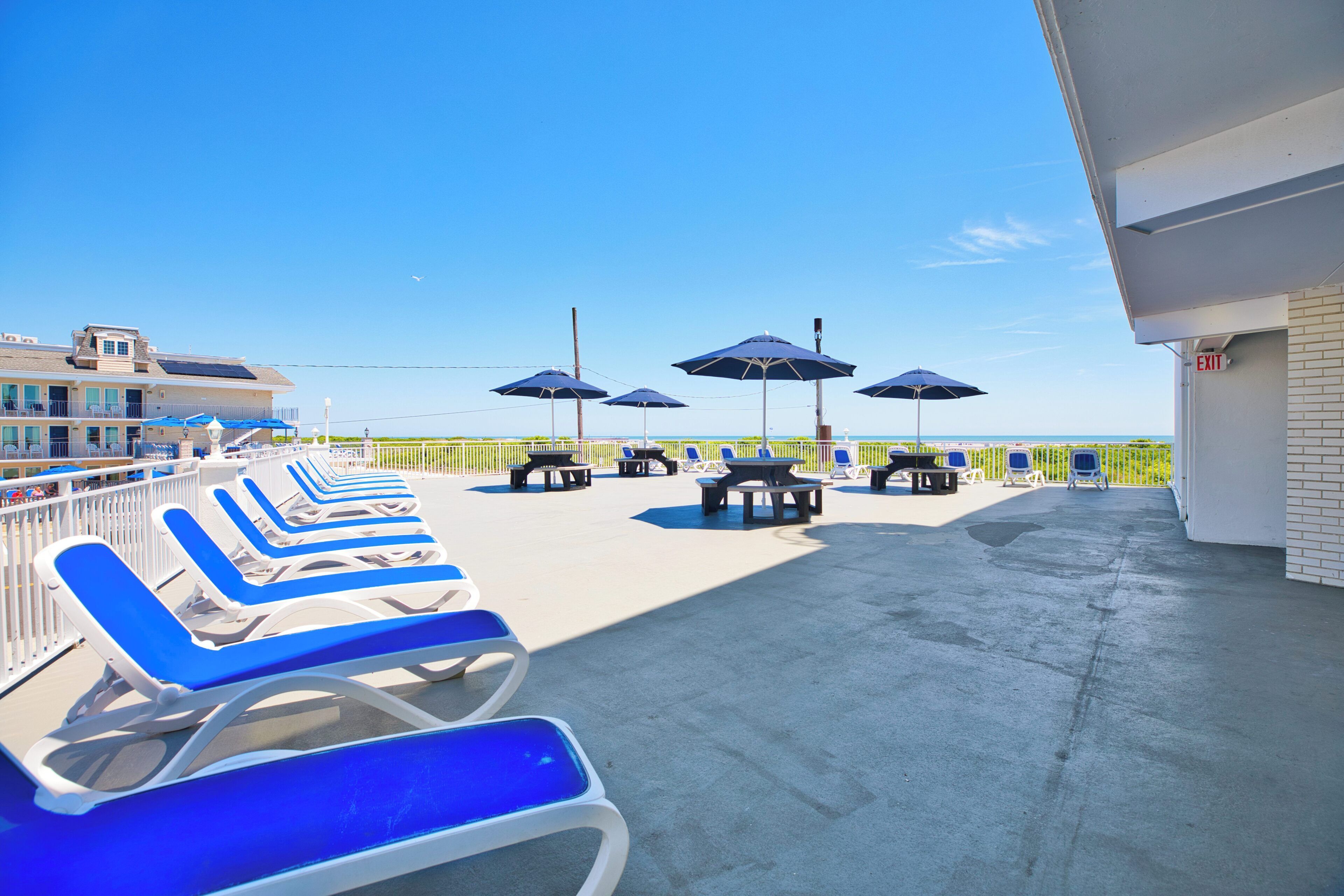 Uma área de piscina no terraço, com espreguiçadeiras e guarda-sóis, um edifício com fachada branca e um céu azul claro.