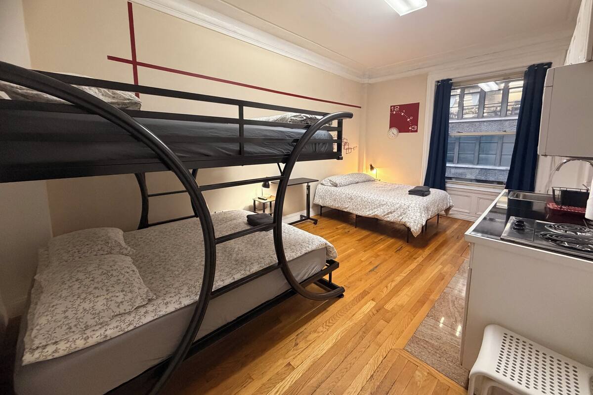 Quarto em dormitório com beliches, uma escrivaninha pequena, um fogão e uma janela com cortinas.