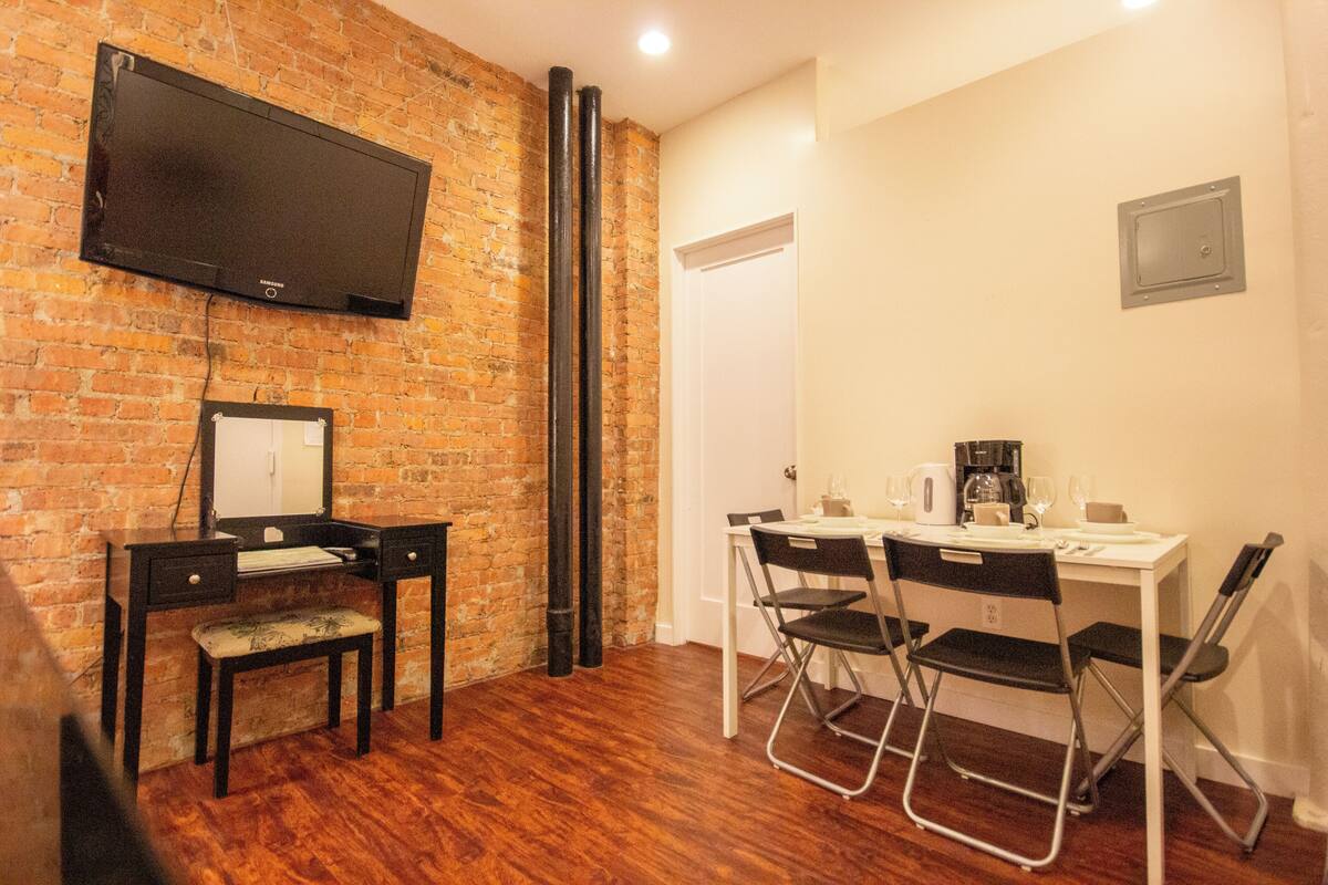 Quarto com parede de tijolos, TV de tela plana fixada acima de uma escrivaninha com um computador, um banco e duas cadeiras.