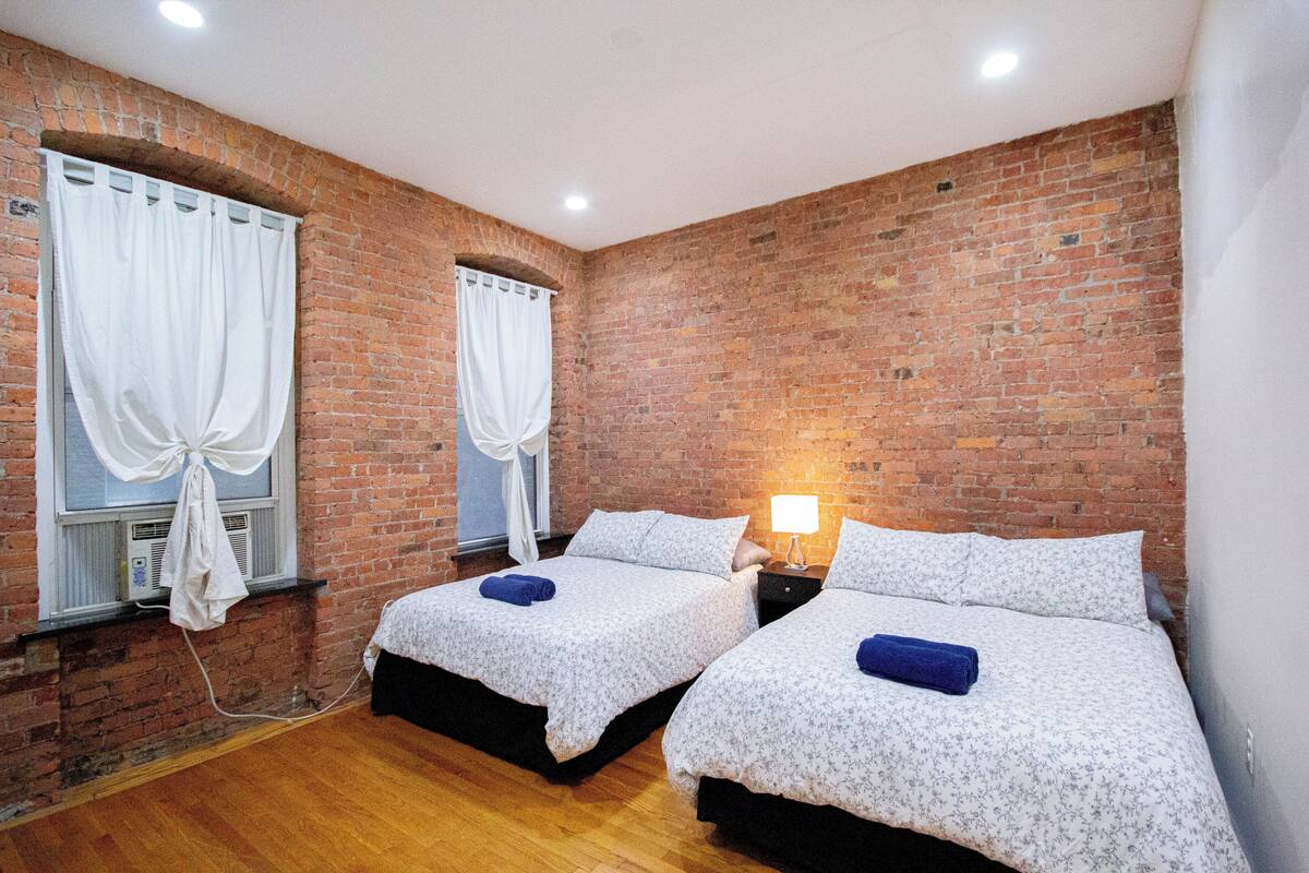 Quarto com duas camas, parede de tijolos e janela com cortinas.