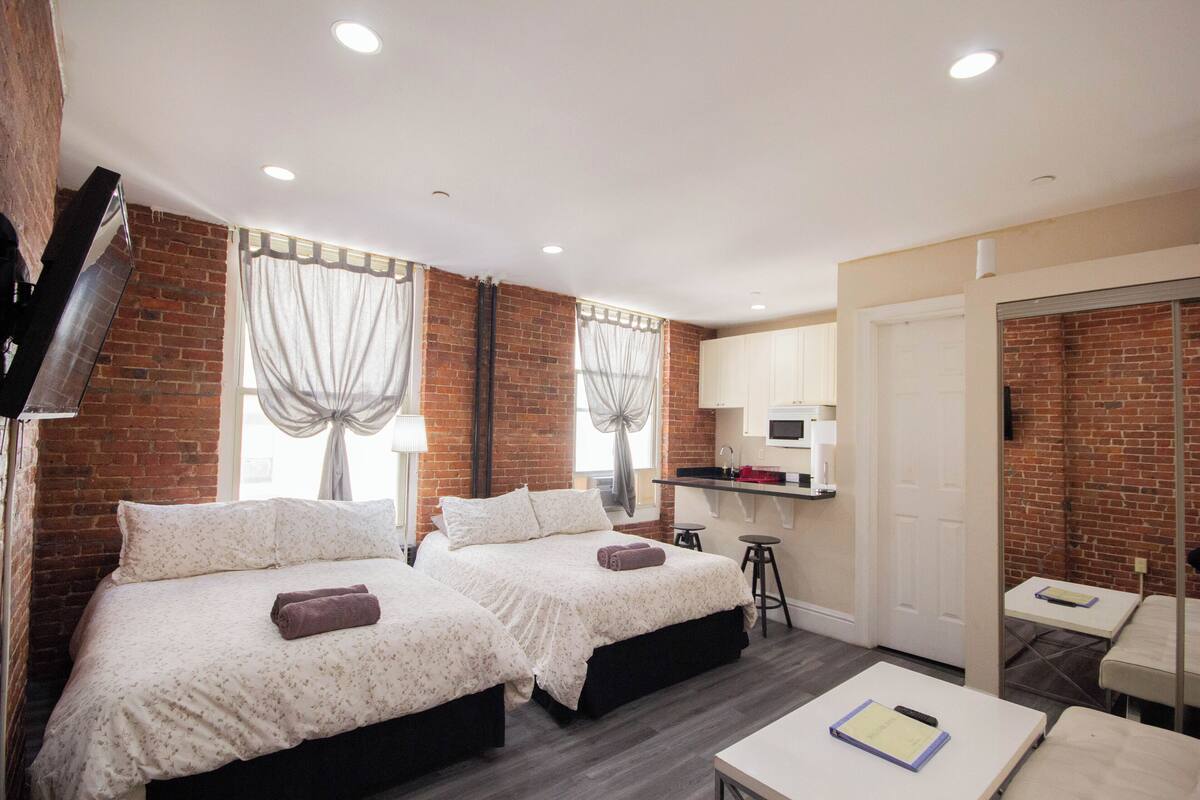 Quarto de hotel com duas camas, uma kitchenette pequena, um espelho e uma parede de tijolos.