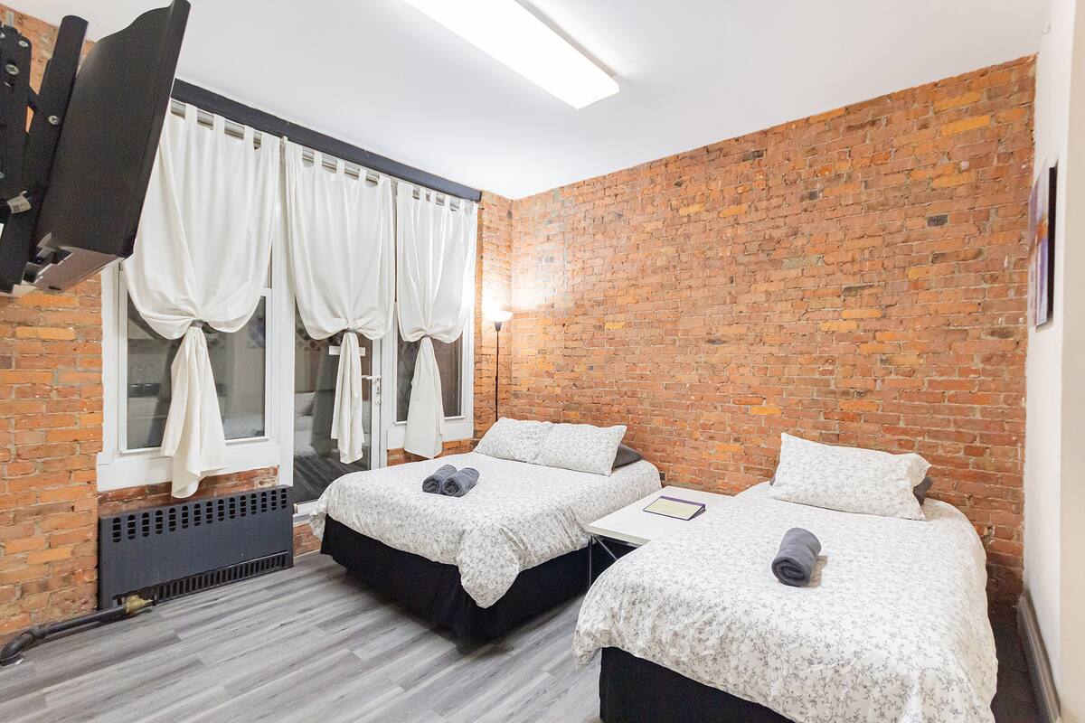 Quarto com duas camas, parede de tijolos, janela com cortinas e televisão de tela plana fixada na parede.