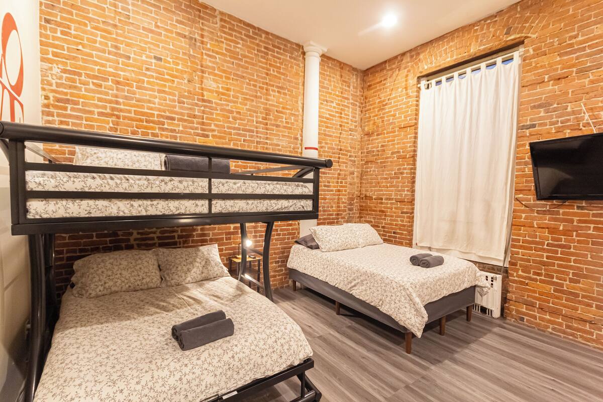 Quarto com duas camas, incluindo uma beliche, paredes de tijolo, televisão de tela plana e janela com cortinas.