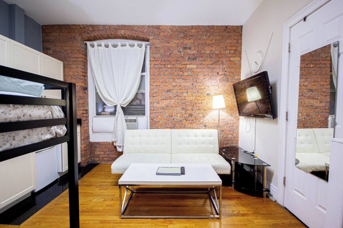 Um quarto com parede de tijolos, sofá e mesa brancos, televisão e uma cama beliche.