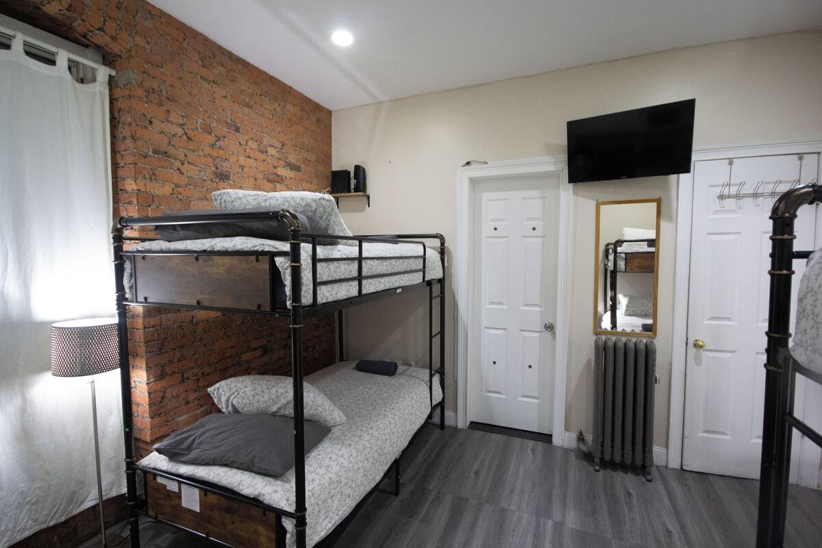 Quarto com beliche, parede de tijolo, porta branca, radiador e TV fixada na parede.