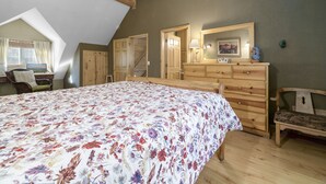 Quarto com uma cama grande, cómoda de madeira e um espelho. Há uma janela com cortinas e uma cadeira.