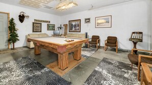 Quarto com mesa de bilhar, dois poltronas, uma cadeira de madeira, uma mesa pequena e uma cabeça de veado presa na parede.