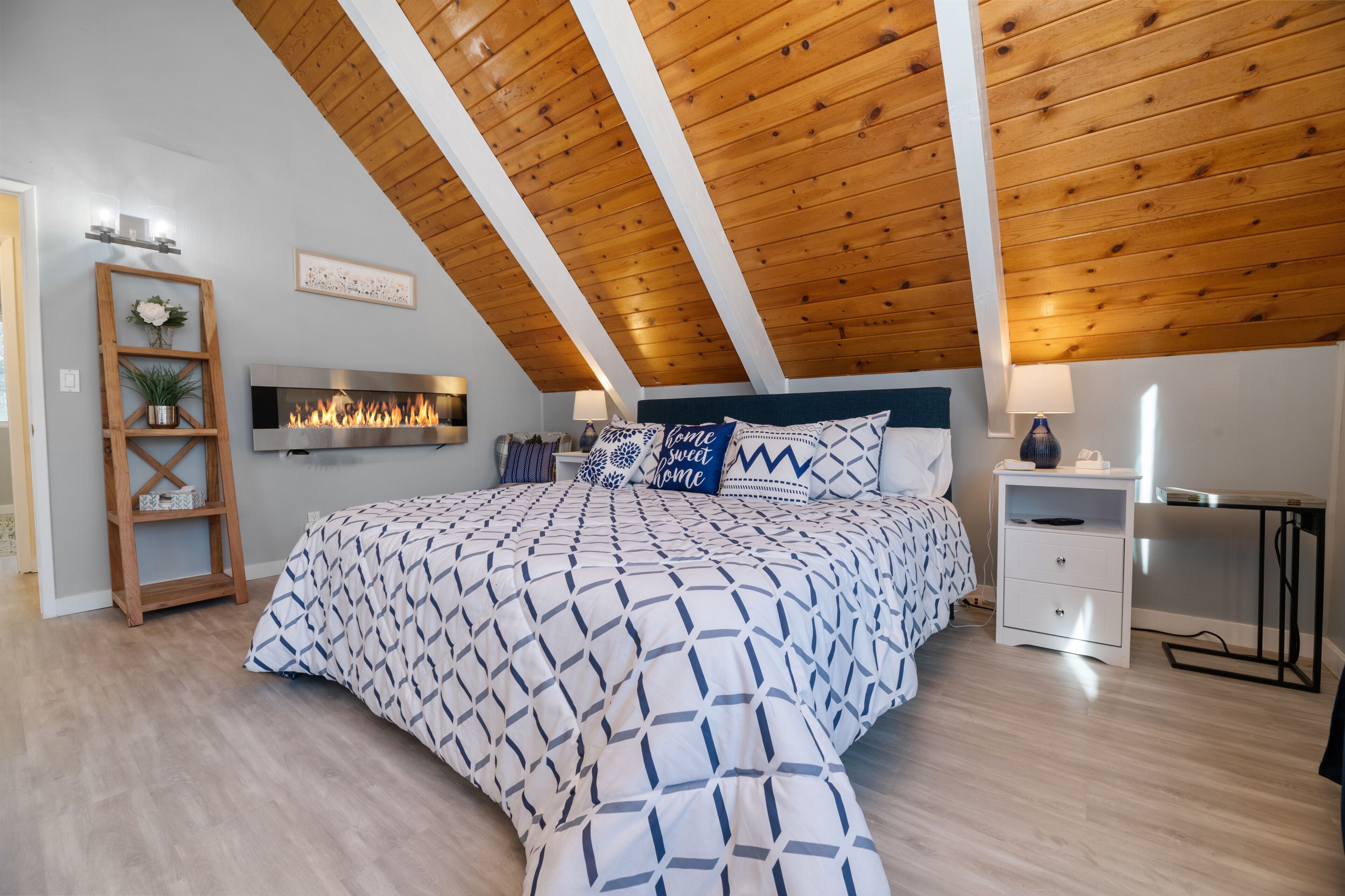 Quarto com teto inclinado, cama com colcha estampada em padrão geométrico, mesas de cabeceira, um criado-mudo e prateleira na parede com uma planta.