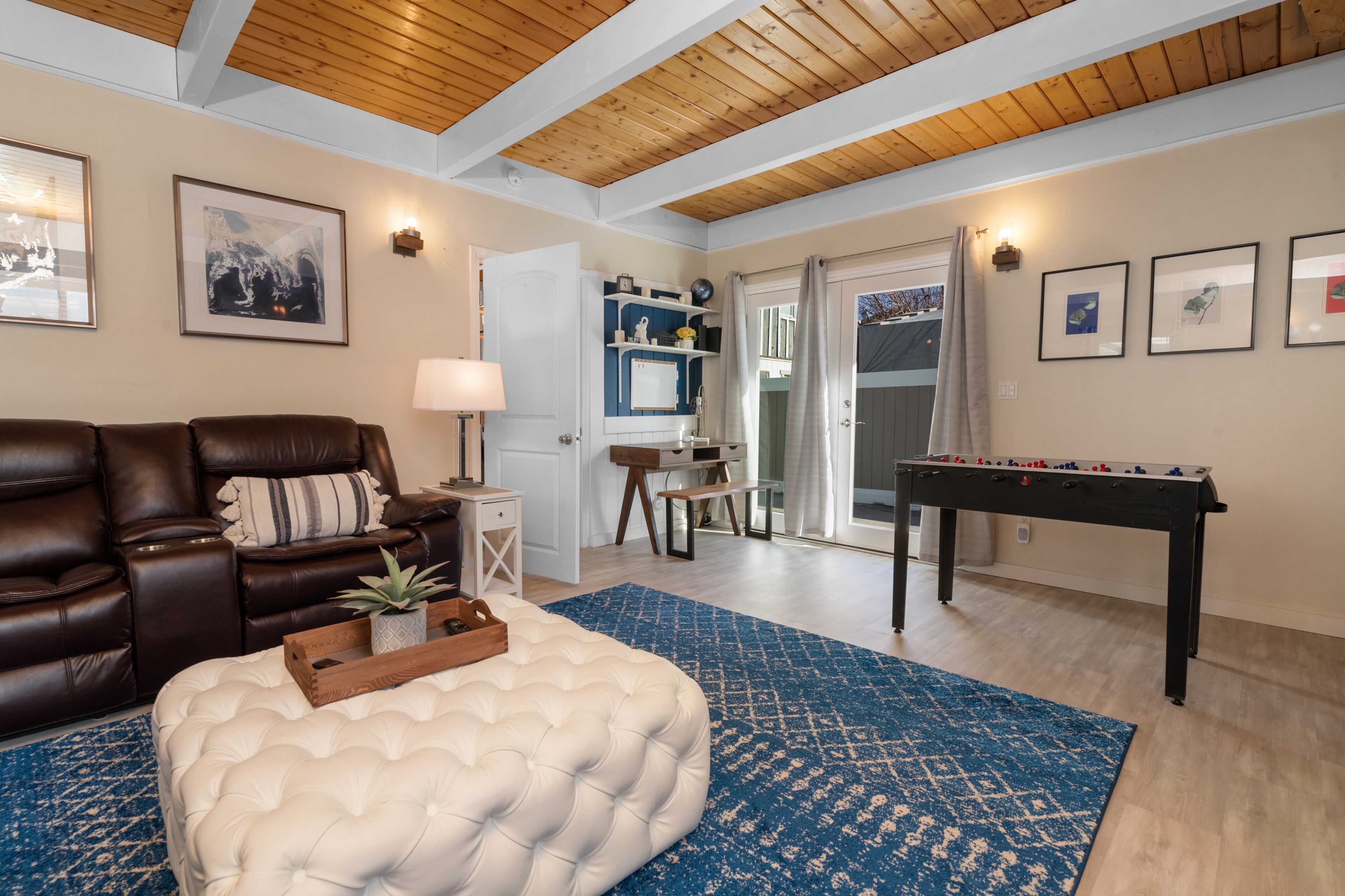 Uma sala de estar com um sofá de couro marrom, um banquinho branco com capitonê, uma mesa de pebolim e um tapete azul estampado.