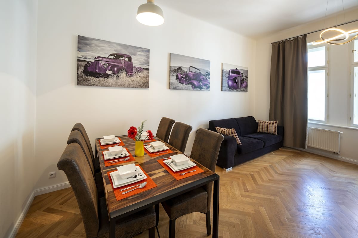 Uma sala de jantar com mesa de madeira posta para quatro pessoas, com dois sofás e uma janela com cortinas.
