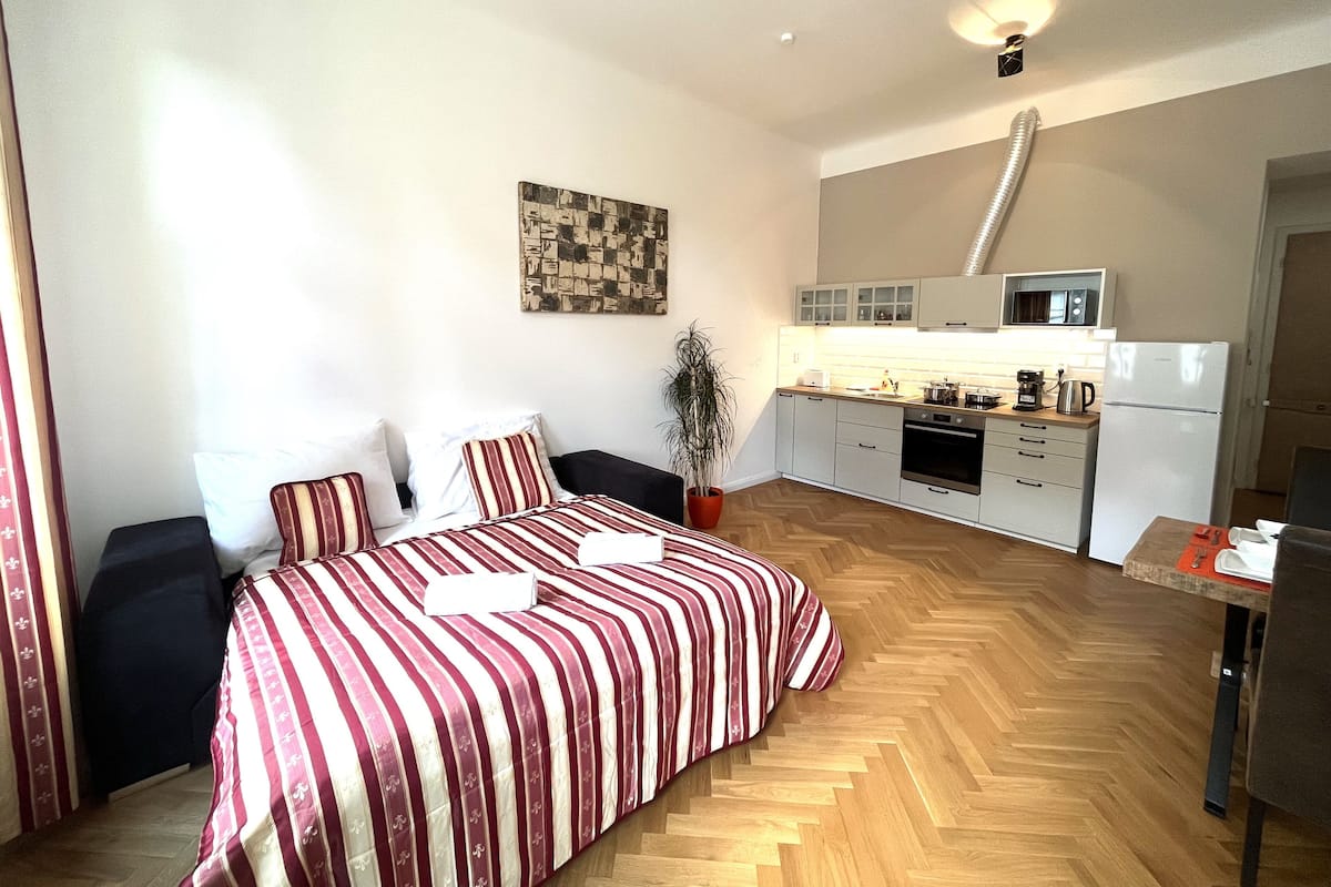 Quarto compacto com cama, mesa de cabeceira e um vaso de planta. Um kitchenette adjacente ao quarto possui micro-ondas, forno e geladeira.