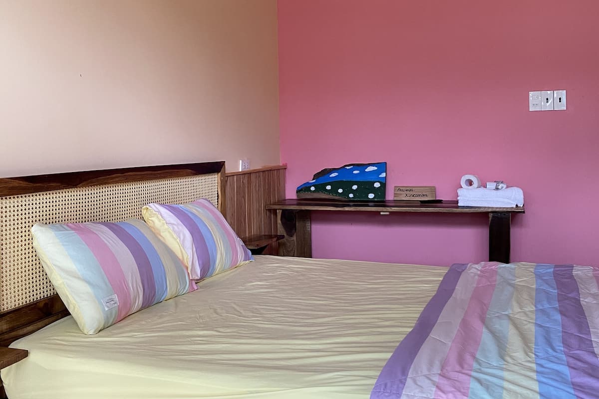 Quarto com estrutura de cama de madeira, travesseiro listrado, parede rosa e uma pequena prateleira com produtos de higiene pessoal.