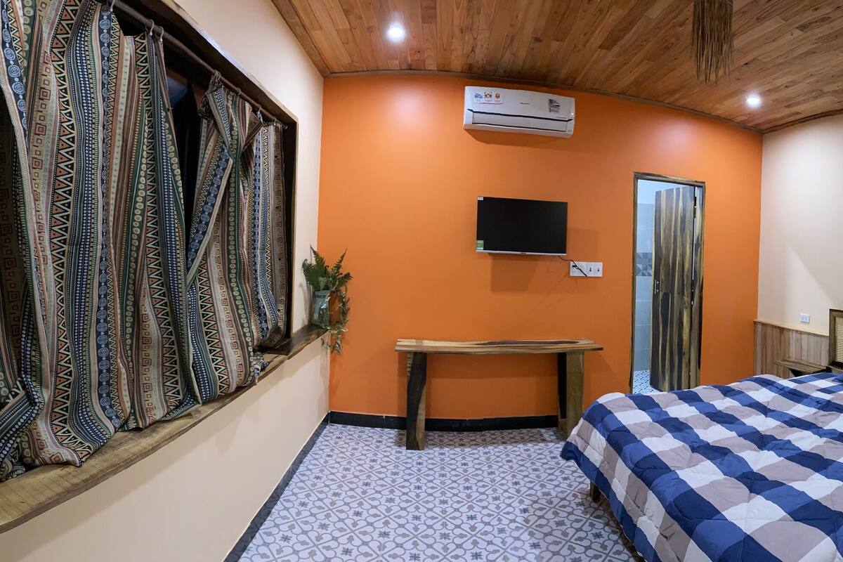 Quarto com colcha xadrez, televisão fixada na parede e janela com cortinas estampadas.