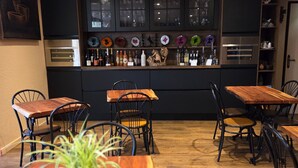 Interior de restaurante com mesas e cadeiras de madeira, área de bar com garrafas e uma estante decorativa com pratos.