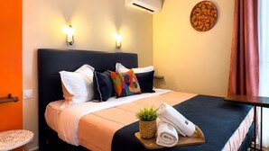 Quarto de hotel com cama, almofadas, mesa de cabeceira, mesa redonda pequena, ar-condicionado fixado na parede e um objeto decorativo na parede.