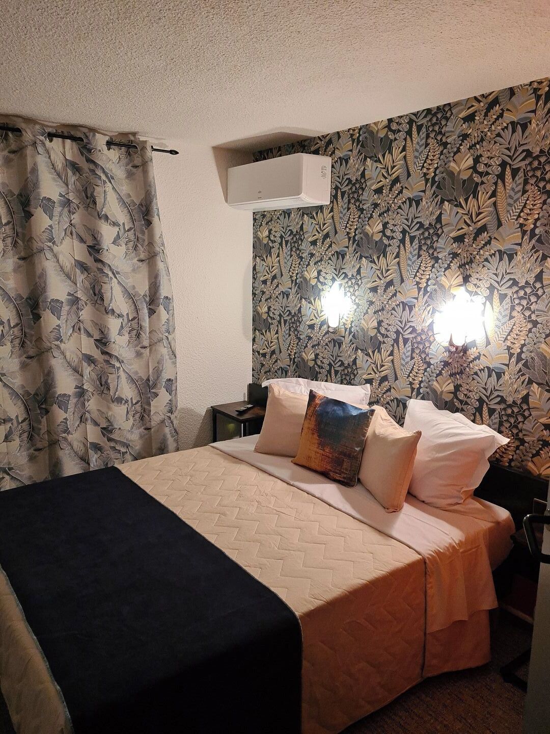 Quarto de hotel com cama, mesa de cabeceira, abajur e parede com padrão de folhas.
