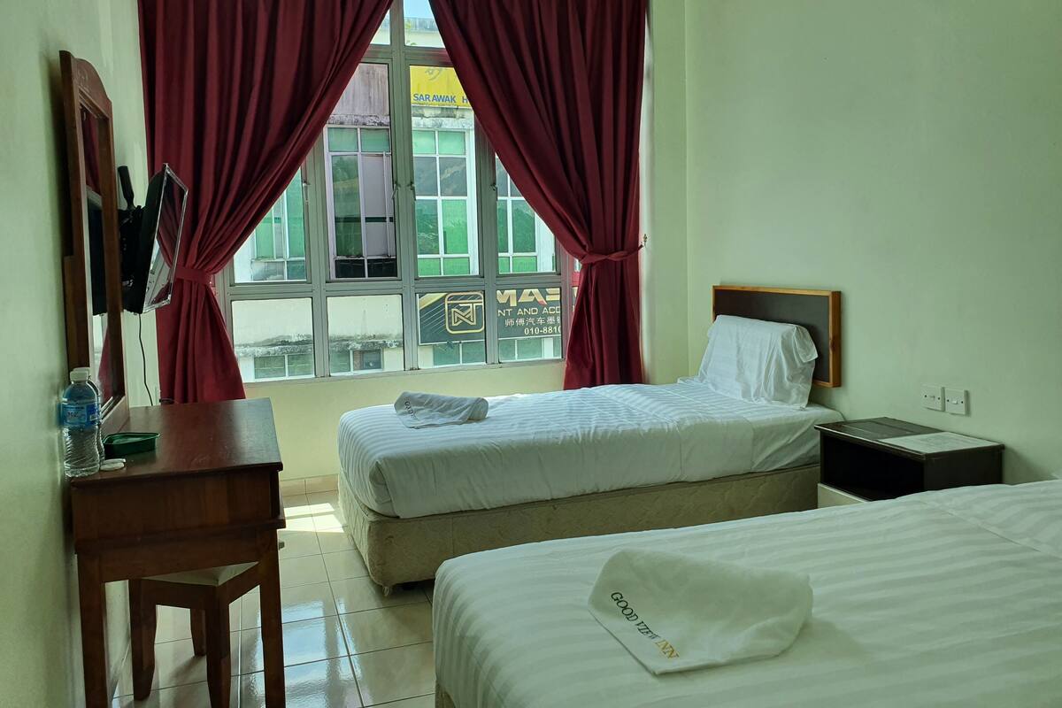 Quarto de hotel com duas camas, uma mesa de trabalho, uma televisão e uma janela com cortinas vermelhas.