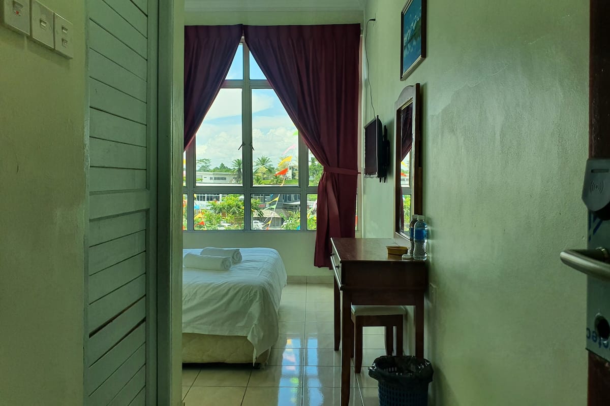 Quarto de hotel com cama, escrivaninha, cadeira, janela com cortinas, televisão fixada na parede e lixeira.
