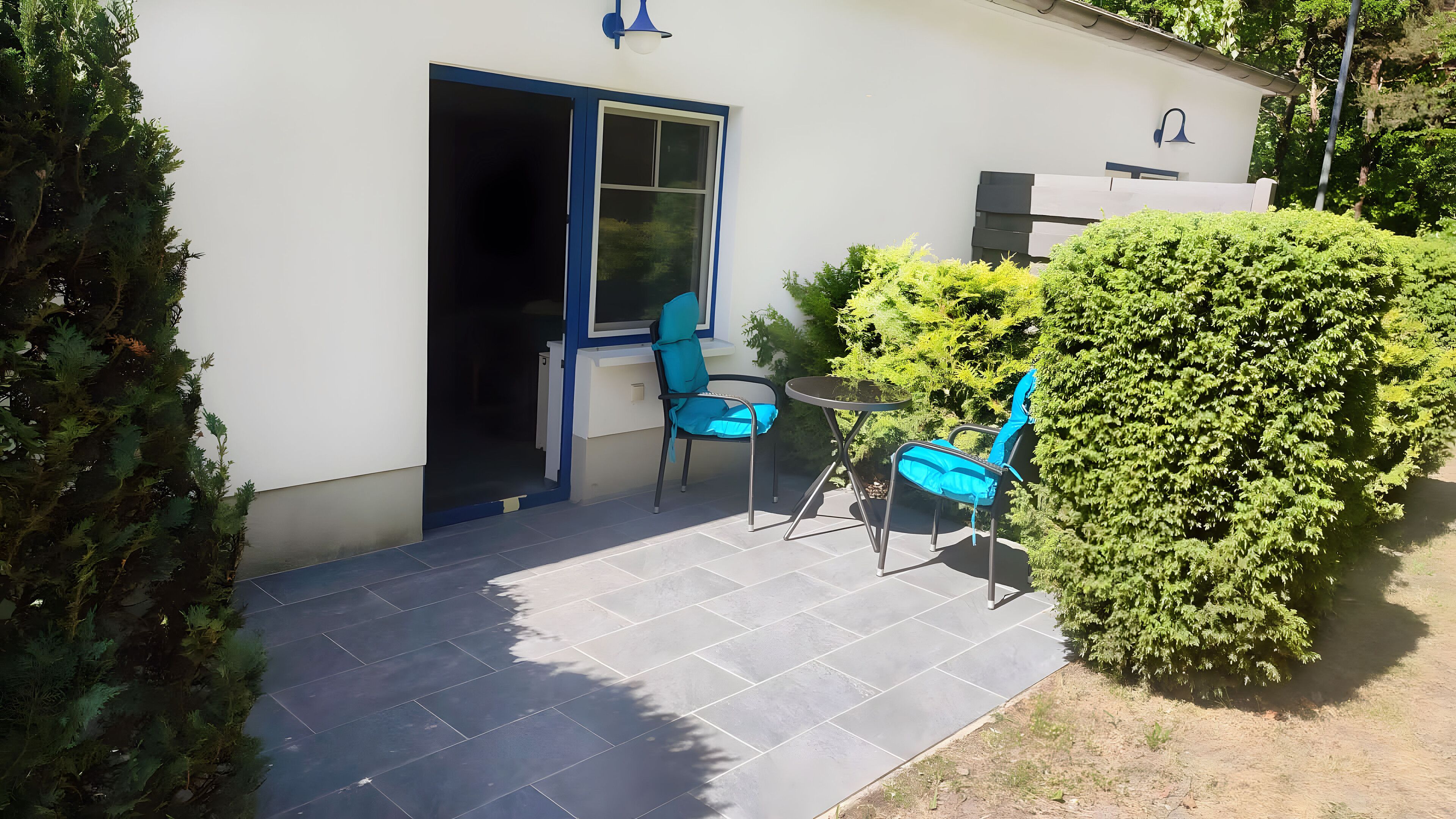 Pátio com duas cadeiras azuis e uma mesa pequena, porta de vidro deslizante e um prédio branco com detalhes em azul.