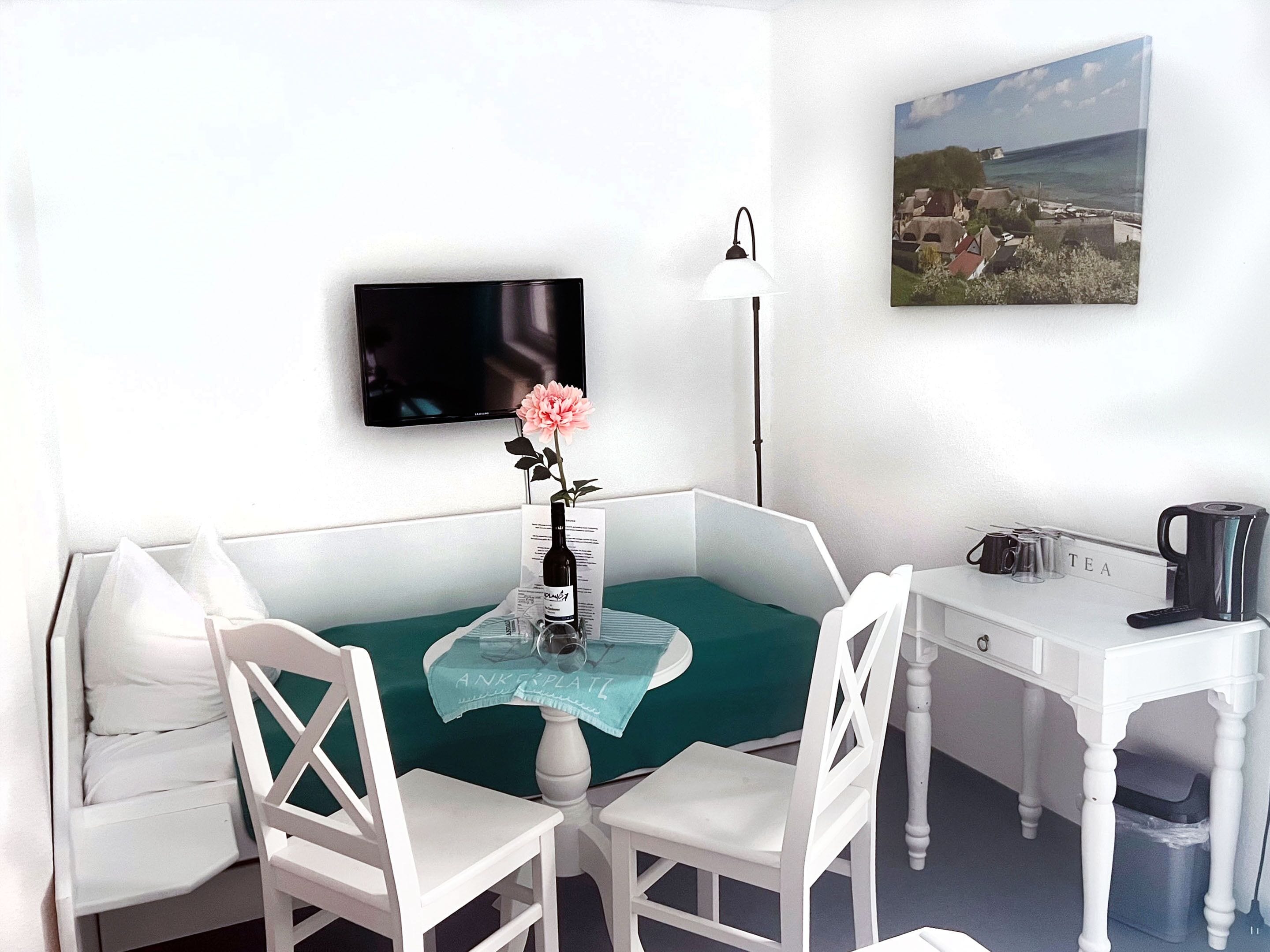 Quarto com sofá branco, uma mesinha com um vaso de flores, televisão na parede, um quadro e uma escrivaninha com chaleira e xícaras.