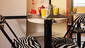 Uma pequena mesa com um abajur vermelho, uma caneca amarela e uma caneca branca. Cadeiras com padrão de zebra estão dispostas ao redor da mesa.