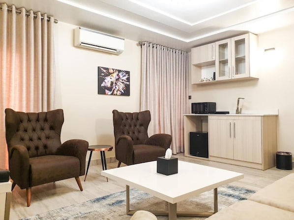 Uma sala de estar moderna com duas poltronas marrons com detalhes em capitonê, uma mesa de centro branca e uma área de cozinha com armários e eletrodomésticos.