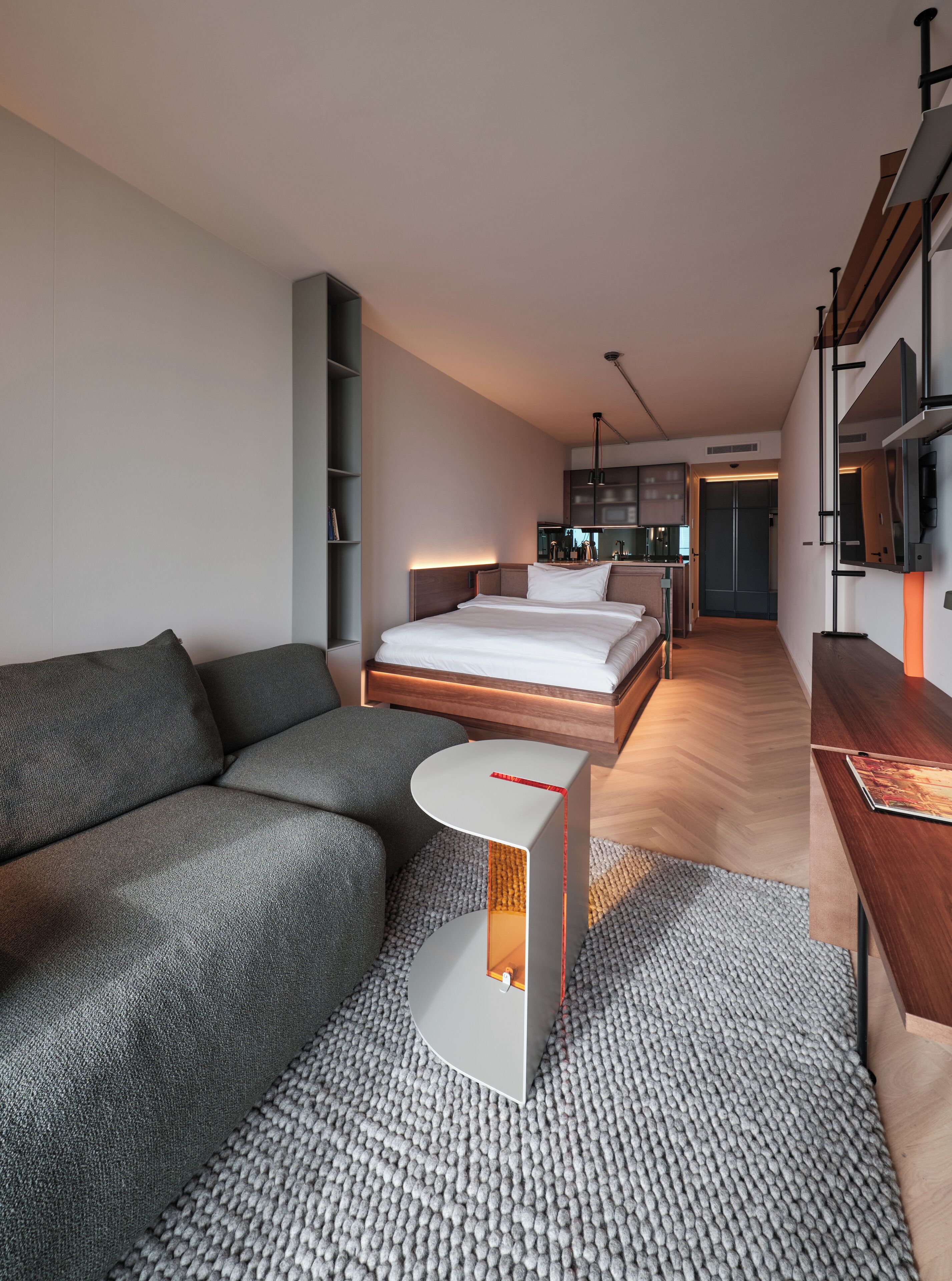 Quarto de hotel moderno com sofá cinza, cama com cabeceira de madeira, mesa lateral branca e piso de madeira em padrão herringbone.
