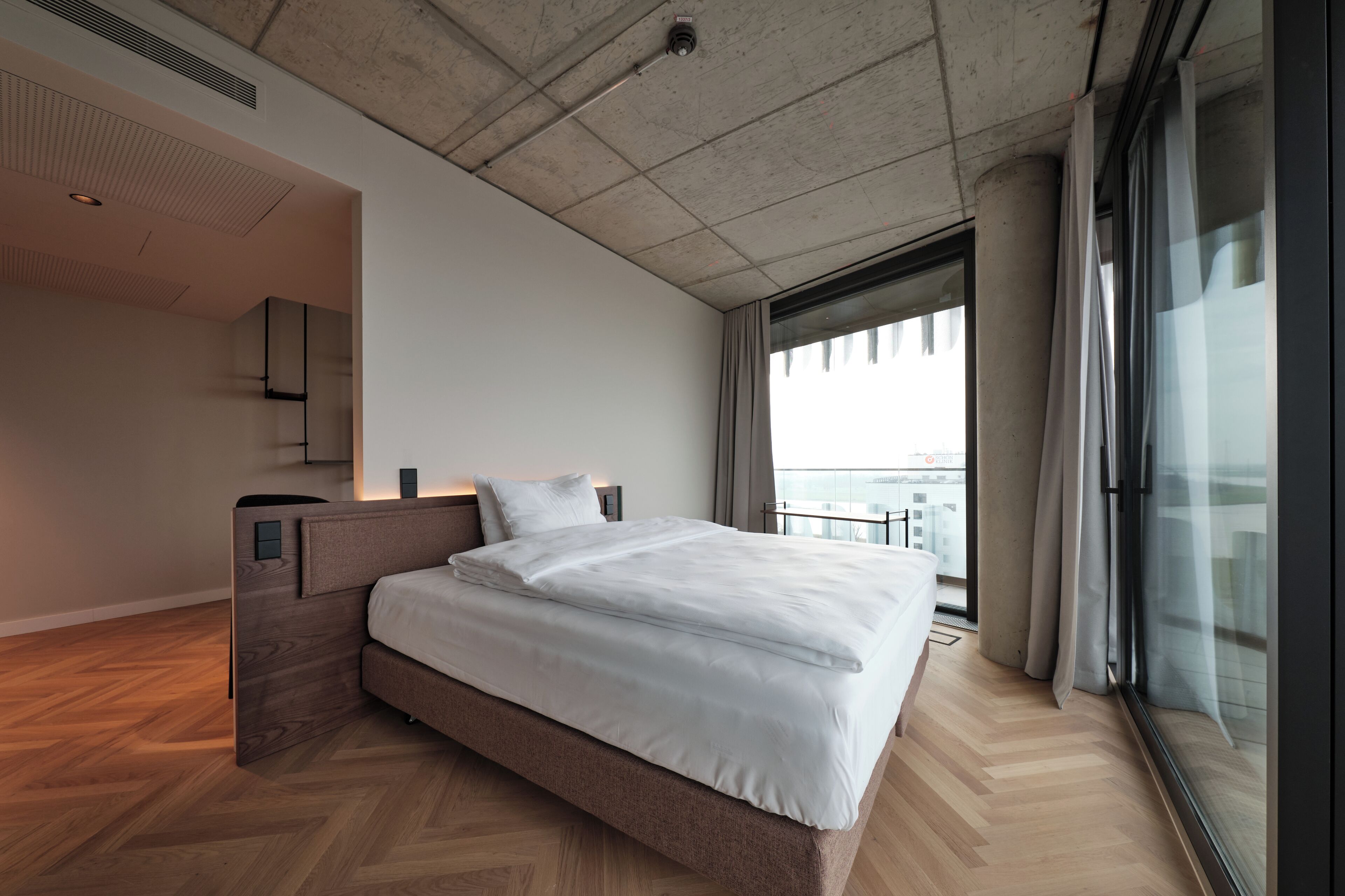 Quarto de hotel moderno com uma cama grande, piso de madeira, um banco embutido e uma janela ampla com vista.