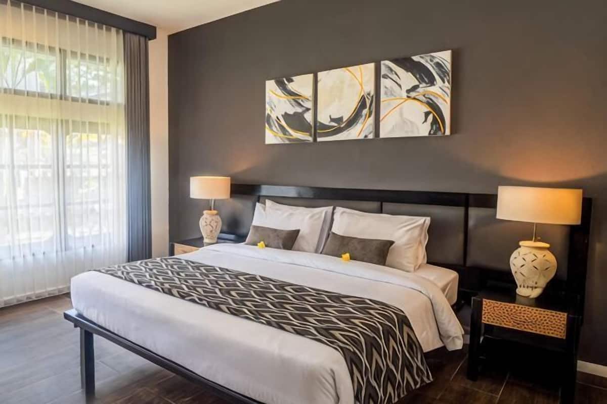 Quarto com cama, duas mesas de cabeceira com luminárias, uma janela grande com cortinas e arte abstrata na parede.