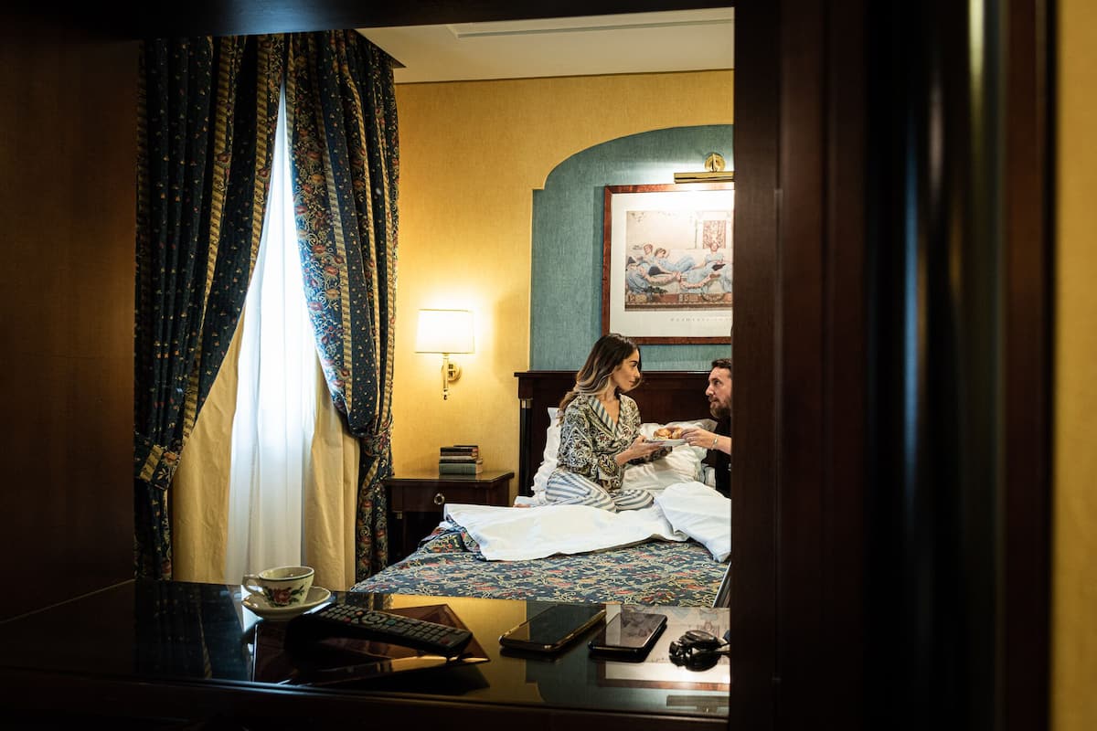 Quarto de hotel com cama, mesa de cabeceira com abajur, um quadro na parede e uma janela com cortinas.