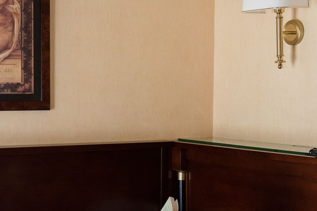 Uma pessoa deitada na cama lendo um livro num quarto com um quadro na parede e uma luminária de cabeceira.