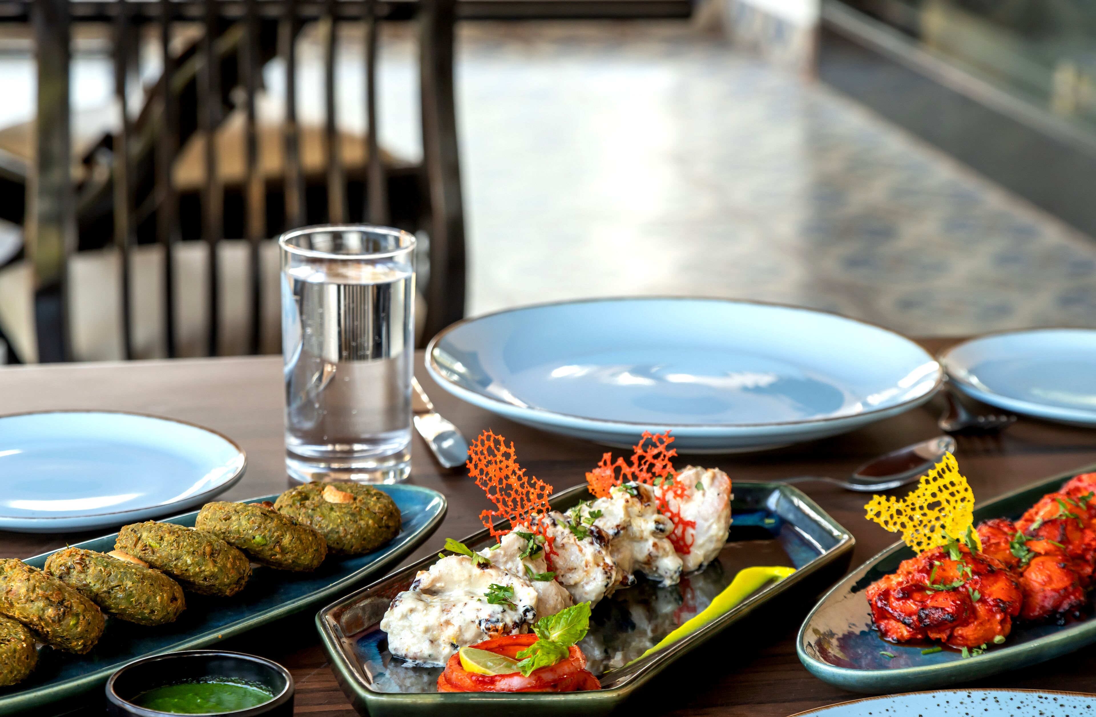 Uma mesa de jantar com diversos pratos, incluindo kebabs, peixe e uma sobremesa, além de um copo de água e pratos.