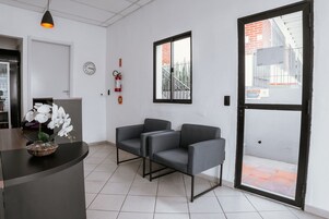 Uma área de recepção com balcão, duas poltronas cinzas, um relógio, um extintor de incêndio e uma janela com vista para um edifício.