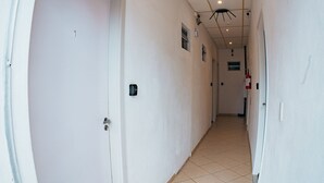 Corredor com paredes brancas, chão em mosaico e portas. Pequenas janelas e luminária no teto.