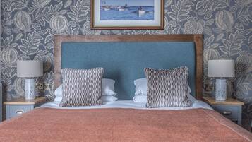 Cama bem arrumada com almofadas estampadas, um quadro acima e duas luminárias de cabeceira em mesas laterais combinando.