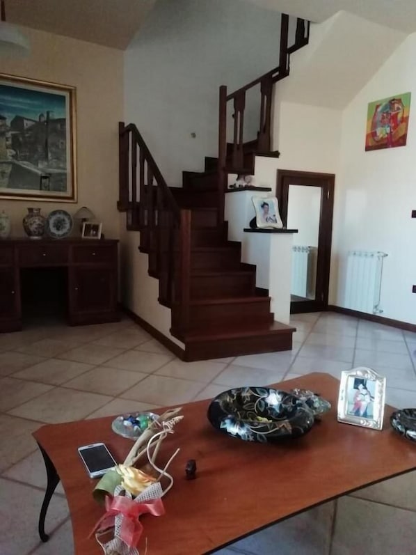 Uma sala de estar ampla com escada de madeira, uma mesa de jantar com um vaso e molduras de quadros, e uma pintura na parede.