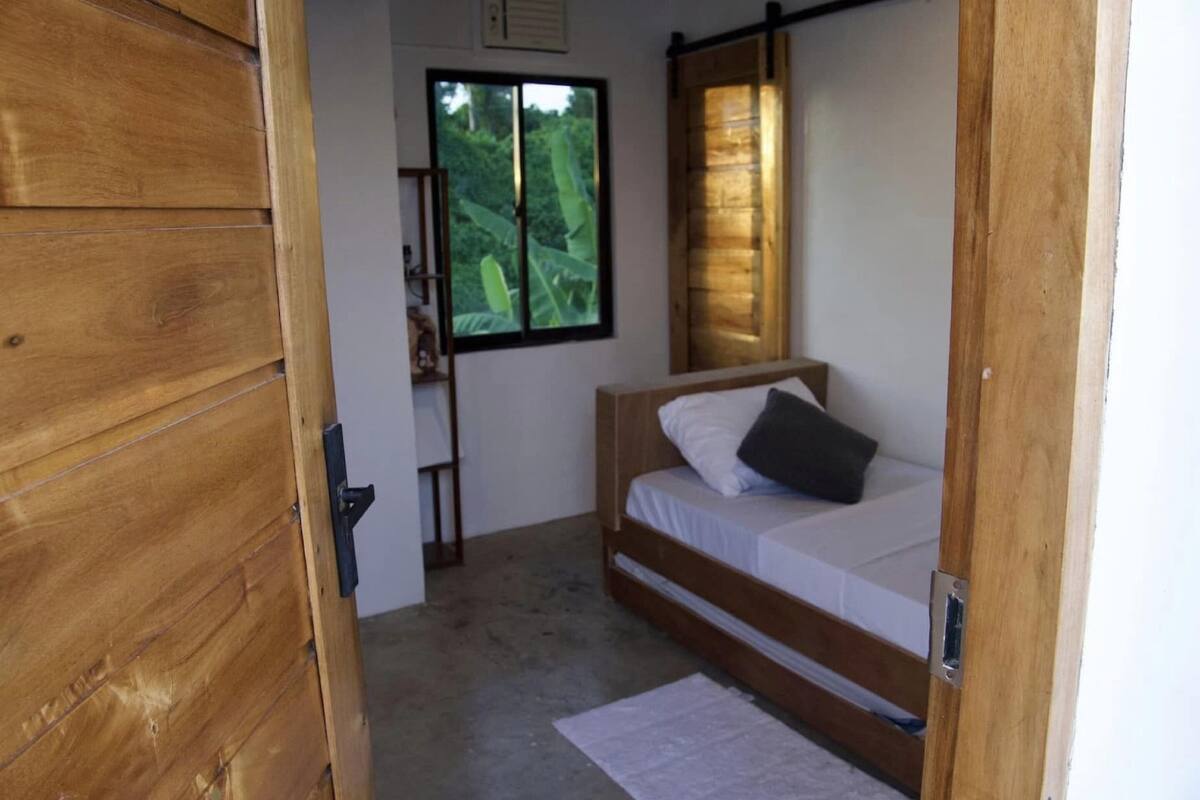 Quarto pequeno, com uma cama de solteiro, porta de madeira, janela com vista para o verde e uma mesinha de cabeceira simples.